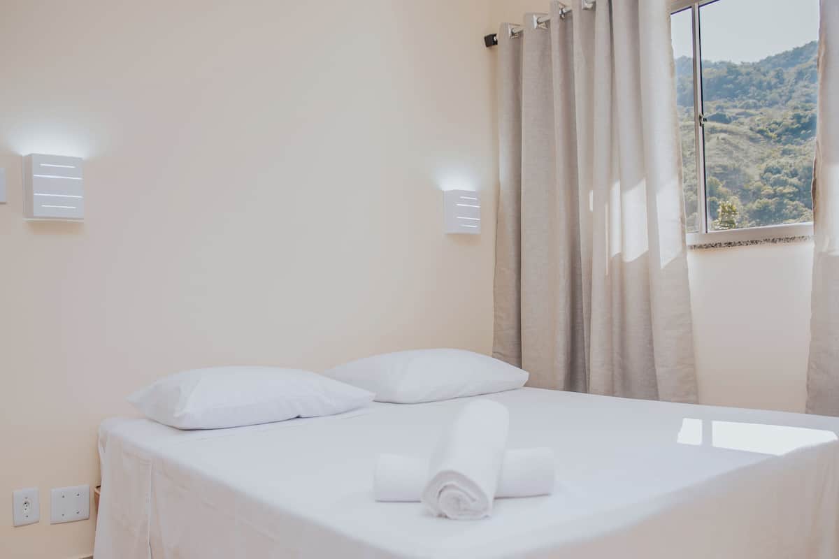 Quarto de hotel com cama, almofadas brancas, toalha enrolada e janela com cortinas.