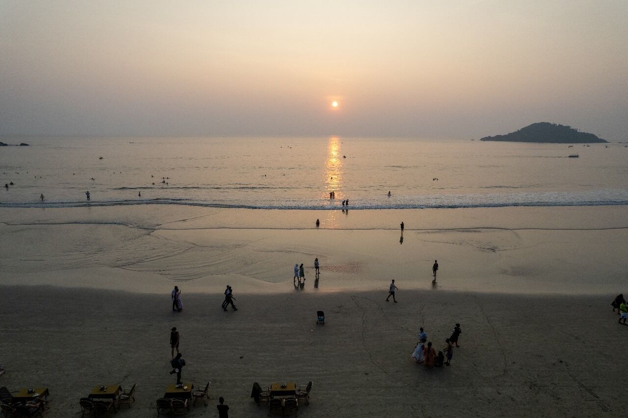 Uma praia com pessoas caminhando, um pôr do sol e uma ilha ao longe.