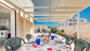 Uma área de refeições ao ar livre com uma mesa posta para dois, coberta por uma pérgula, e vista para os edifícios ao redor.