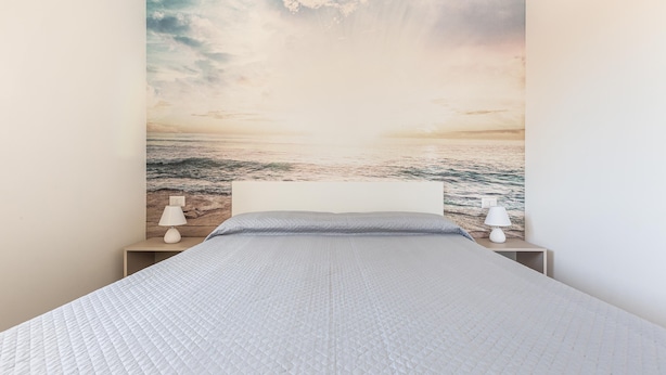 Quarto com cama, duas mesas de cabeceira com luminárias e um mural na parede retratando uma paisagem praiana.