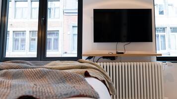 Quarto de hotel com cama, televisão na parede, radiador e janela com vista para edifícios.