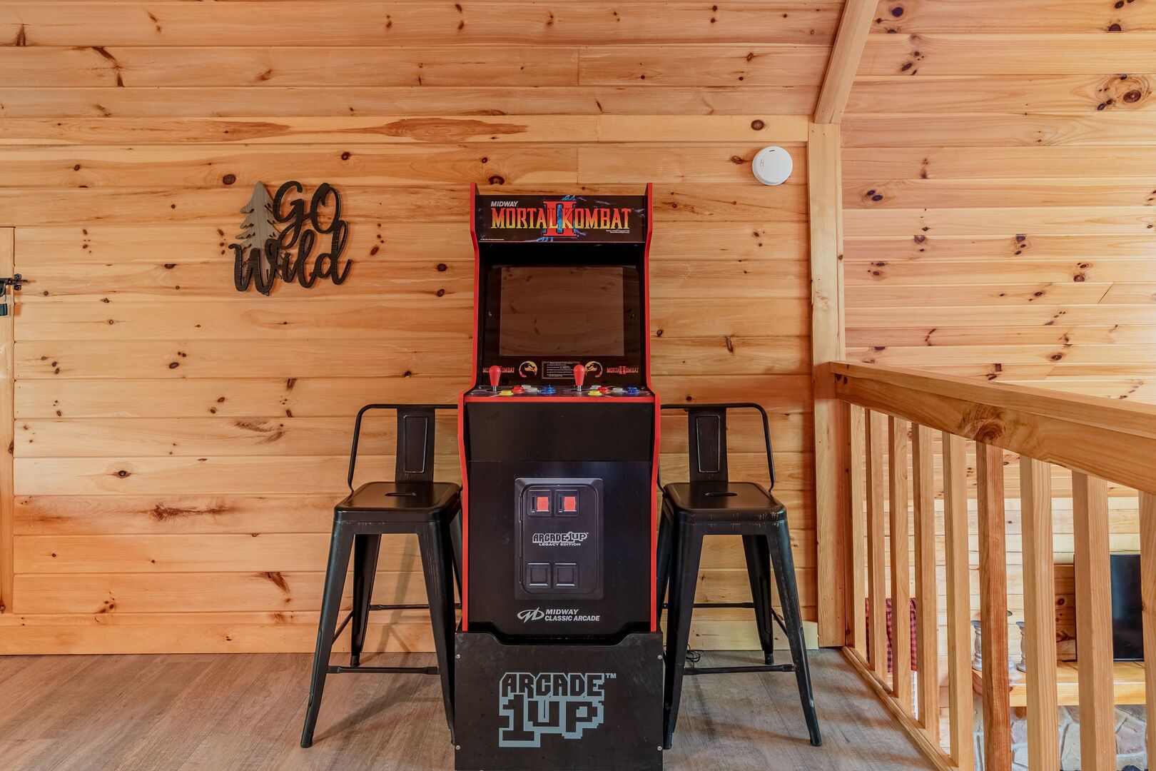 Interior de uma cabana de madeira com um fliperama Midway com o nome 'Mortal Kombat' e um console 'Arcade 1UP'.