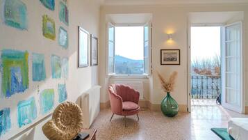 Um quarto espaçoso com uma grande janela, uma cadeira rosa, um vaso verde e uma parede com pinturas abstratas.