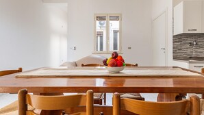 Uma área de refeições com uma mesa de madeira, cadeiras, uma tigela de frutas e uma janela com persianas.