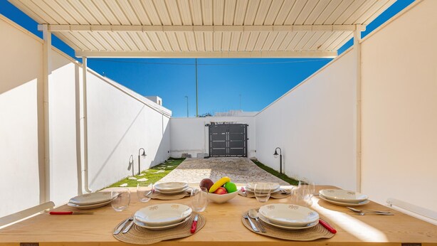 Mesa de jantar posta para quatro pessoas, com uma tigela de frutas no centro, sob um teto branco com detalhes azuis.