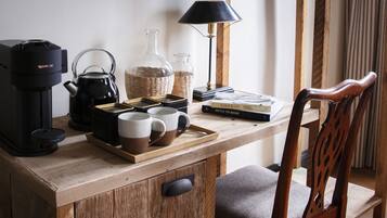 Uma mesa de madeira com uma cafeteira, canecas, um tabuleiro, uma luminária e livros.