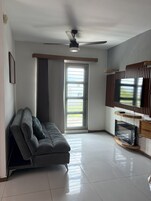 Uma sala de estar moderna com um sofá cinzento, um ventilador de teto, uma janela grande e uma televisão de tela plana fixada em uma parede de madeira.
