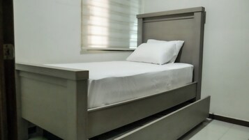 Uma cama de solteiro com colchão e travesseiro brancos, cabeceira de madeira e janela com persianas.