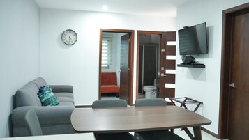 Uma sala de estar moderna com um sofá cinzento, uma mesa de jantar de madeira e uma TV fixada na parede.