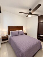 Quarto com cama, mesas de cabeceira, ventoinha no teto e abajur.