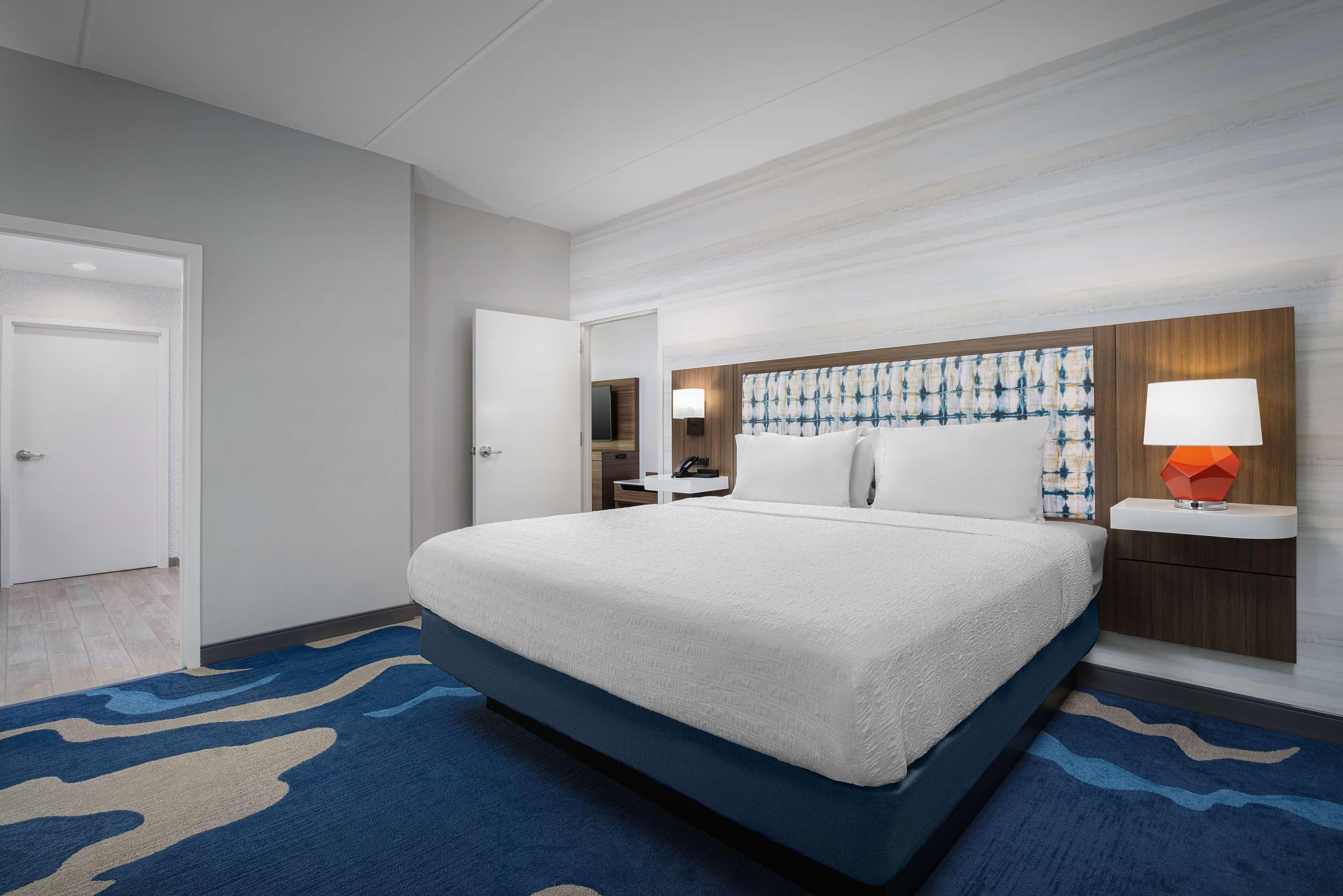 Quarto de hotel moderno, com uma cama grande, uma mesa de cabeceira e uma luminária, e um banheiro visível por uma porta aberta.