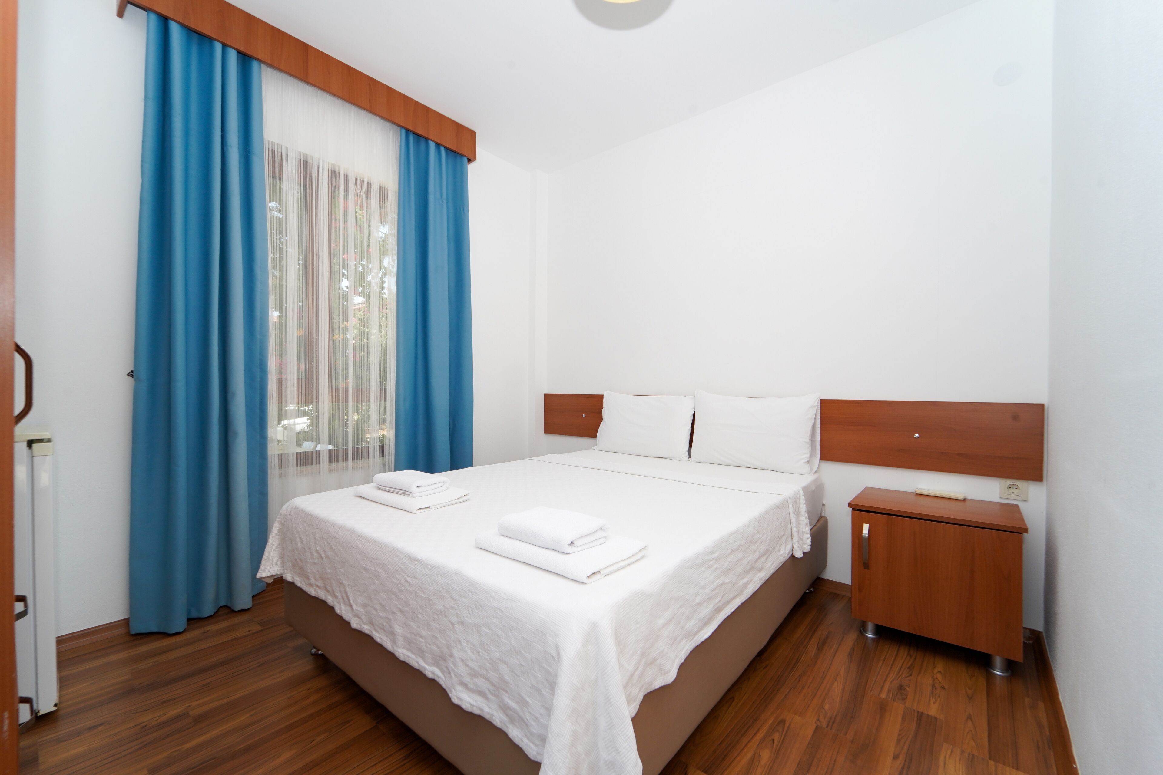 Quarto de hotel limpo e moderno, com uma cama grande, cabeceira de madeira, mesa de cabeceira pequena de madeira e uma janela com cortinas azuis.
