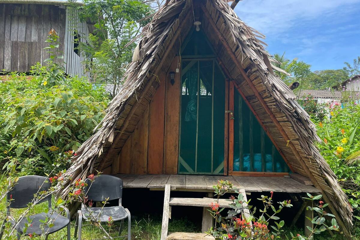 Uma cabana com telhado de palha, porta verde e pequena varanda com duas cadeiras.