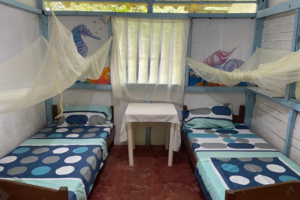 Um quarto pequeno e fechado com duas camas, uma mesa e uma janela com cortinas.