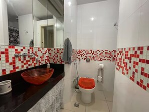 Casa de banho com bancada preta, lavatório vermelho, sanita branca com tampa laranja e área de chuveiro.