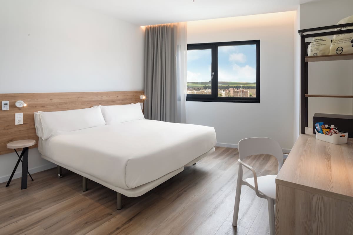 Quarto de hotel com cama, mesa de cabeceira, cadeira e janela com vista para a cidade.