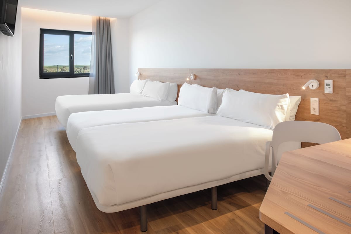 Quarto de hotel com duas camas, cabeceira de madeira, escrivaninha e janela com vista.