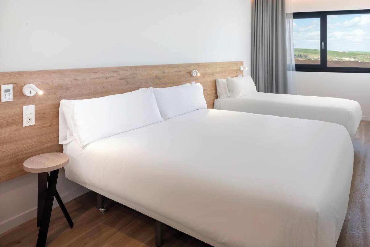 Quarto de hotel com duas camas, uma mesa redonda pequena e uma janela com vista para o campo.