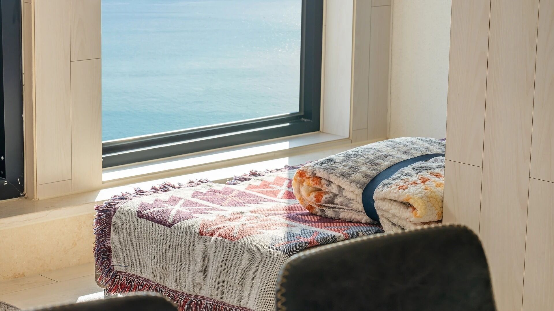 Quarto com vista para o mar através de uma janela, uma cama com manta estampada e uma cadeira.