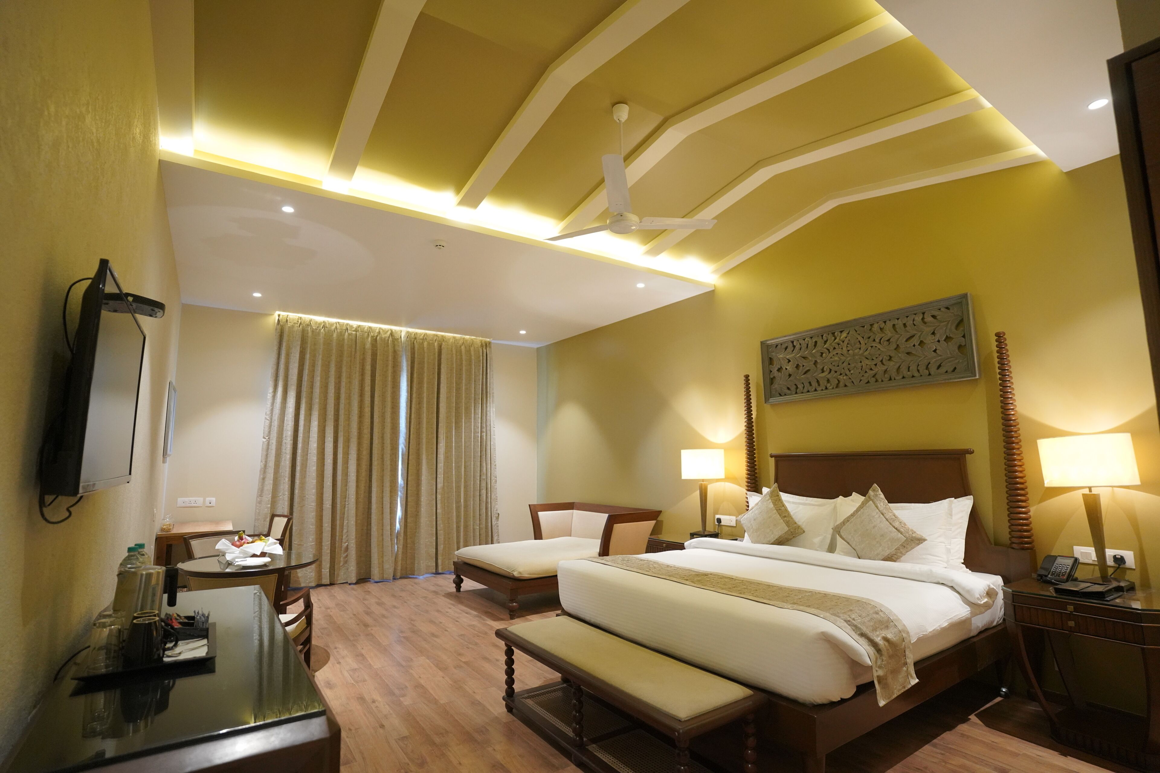Quarto de hotel com uma cama grande, mesas de cabeceira, escrivaninha, cadeira, banco, televisão e cortinas.