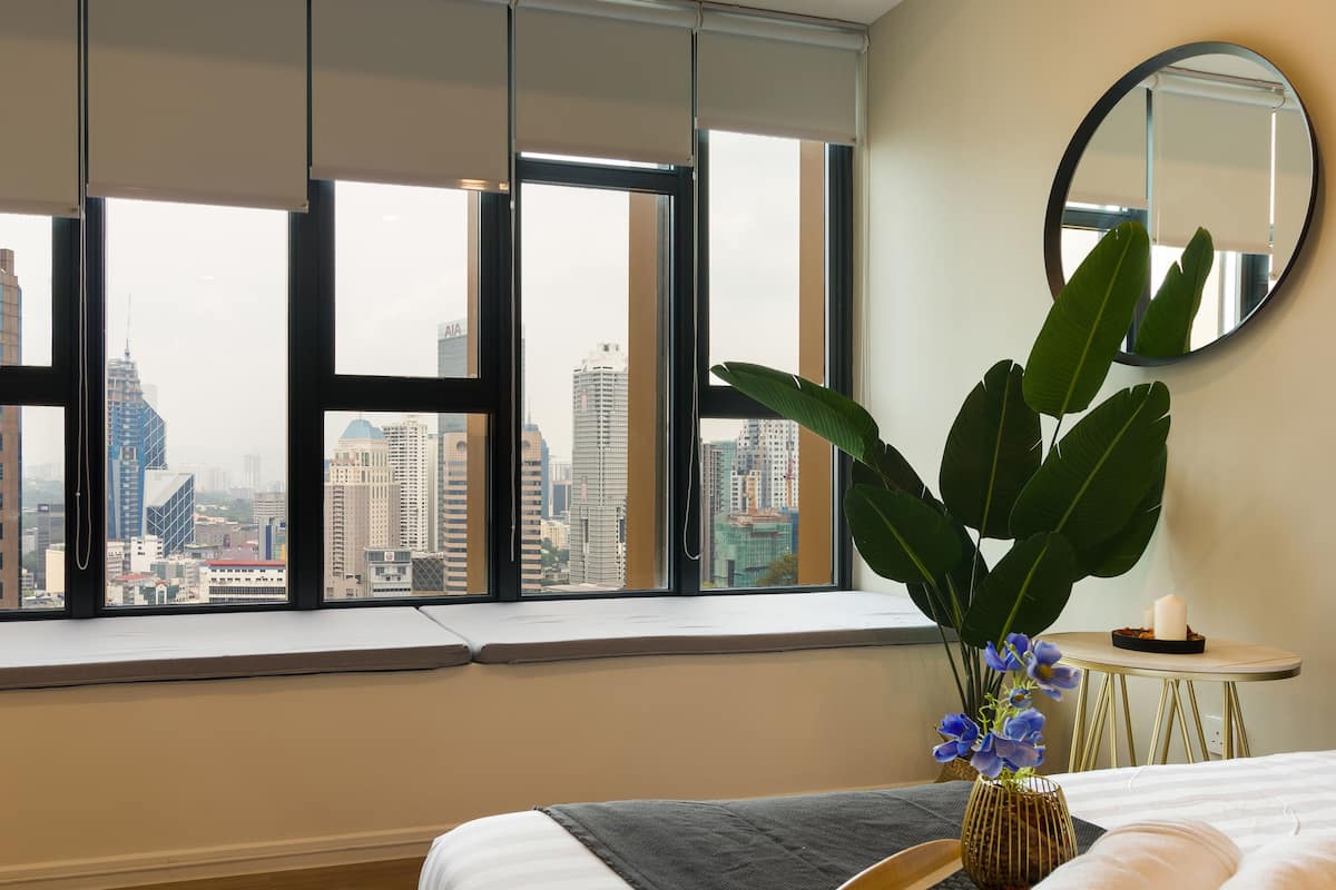 Quarto de hotel com uma janela grande que oferece vista para a cidade, uma cama com roupa de cama branca, uma mesa redonda pequena com uma vela e uma planta em vaso.
