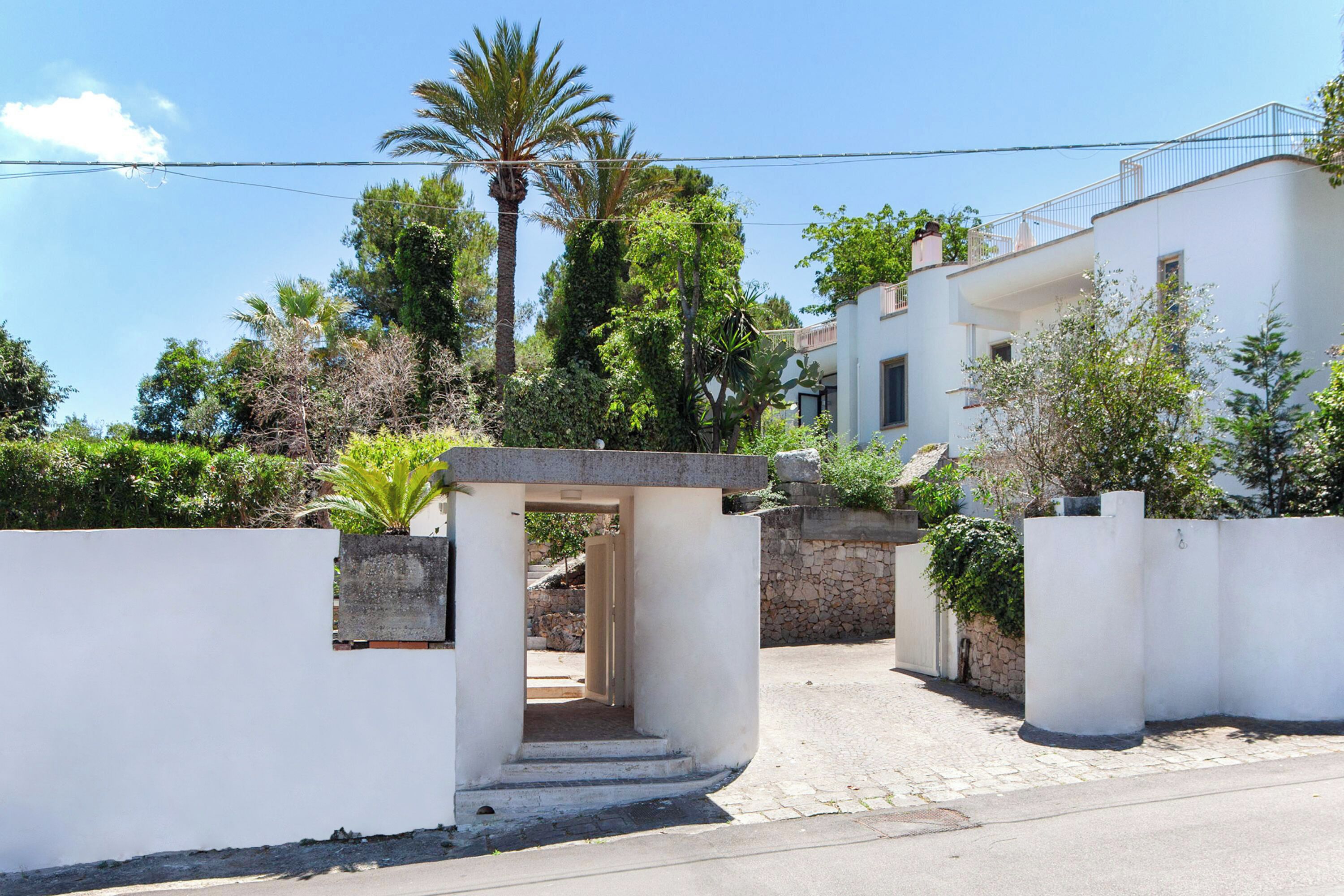 Uma entrada protegida por grades para um edifício, com um caminho de pedra que leva à porta, rodeada por vegetação e um céu claro.