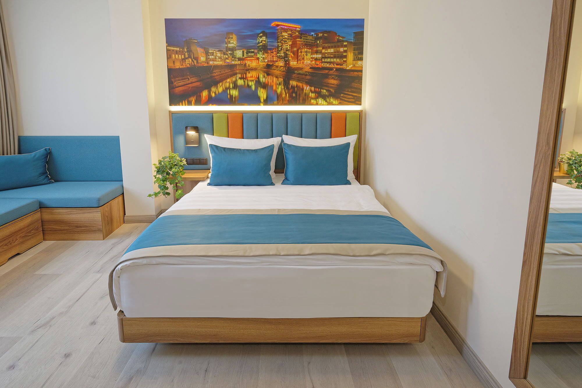 Quarto moderno com cama grande, pintura de paisagem urbana acima da cama e sofá azul no canto.
