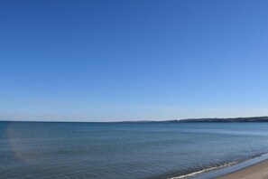 Vista nítida de um mar calmo, com uma praia arenosa, terra à distância e um céu limpo.