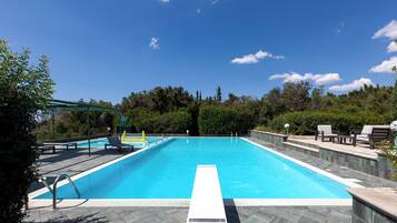 Piscina retangular com plataforma de mergulho ao centro, ladeada por azulejos de pedra e equipada com espreguiçadeiras e uma pequena área de deck.