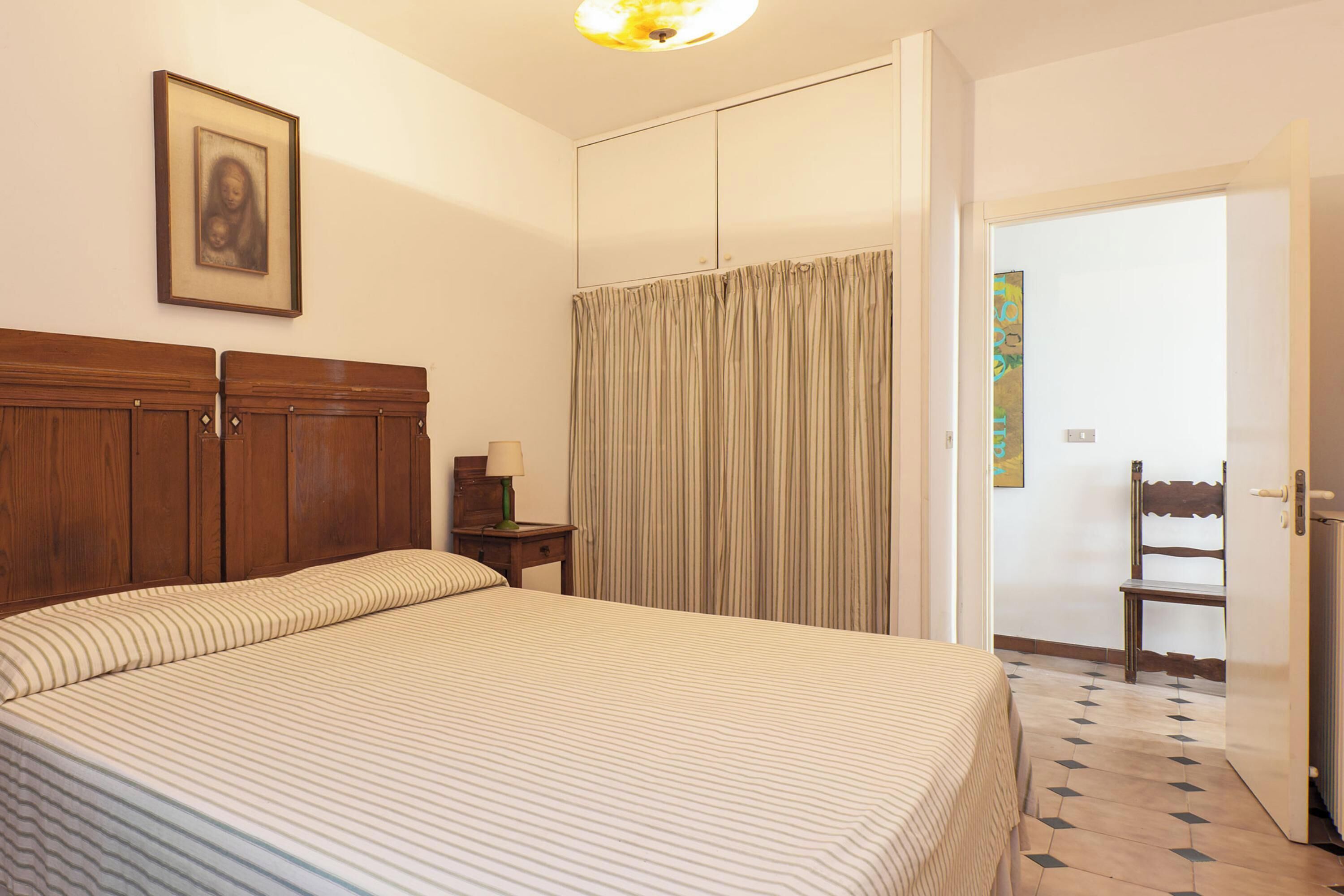 Quarto com uma cama grande, cabeceira de madeira, mesa de cabeceira com abajur, quadro na parede, cortina e porta que dá para outro cômodo.