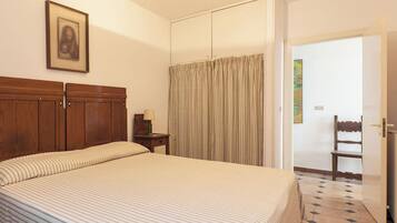 Quarto com uma cama grande, cabeceira de madeira, mesa de cabeceira com abajur, quadro na parede, cortina e porta que dá para outro cômodo.
