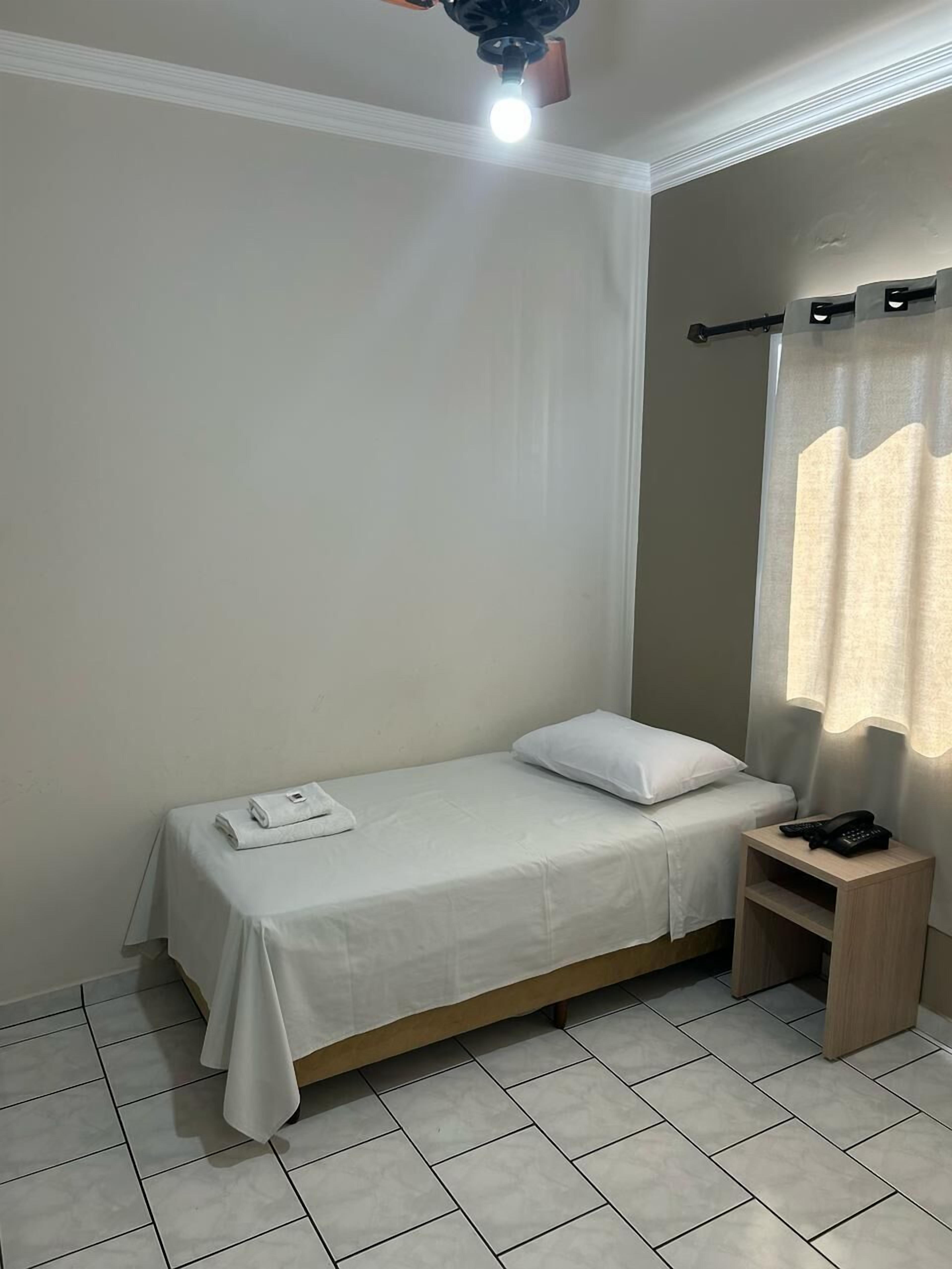 Uma cama pequena e bem arrumada com lençol e travesseiro brancos, uma mesinha de cabeceira de madeira com um telefone e um ventilador de teto.