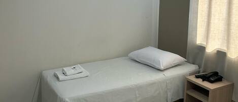 Uma cama pequena e bem arrumada com lençol e travesseiro brancos, uma mesinha de cabeceira de madeira com um telefone e um ventilador de teto.