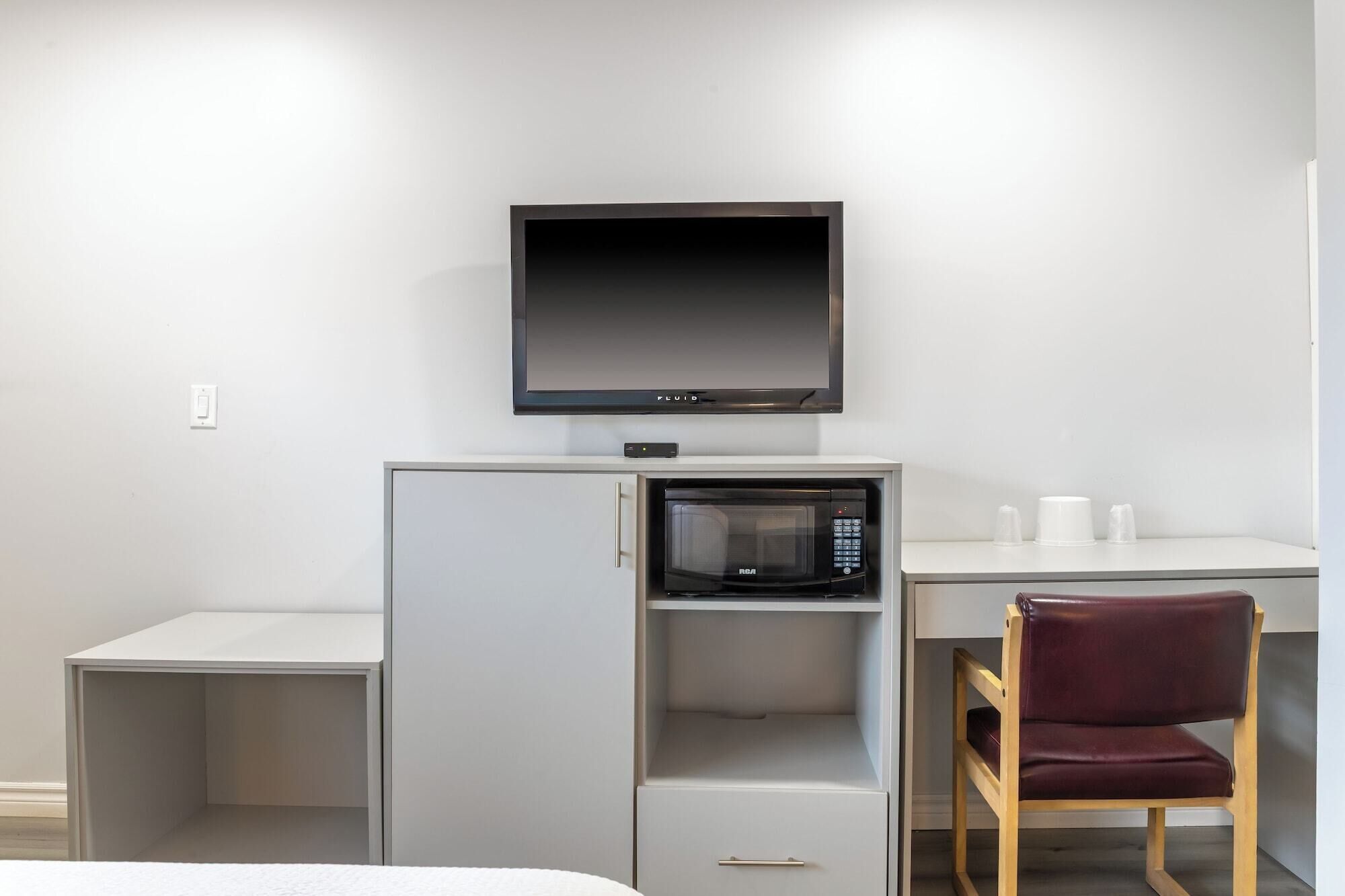 Quarto compacto com televisão de tela plana montada acima de um micro-ondas embutido, uma pequena escrivaninha com cadeira e uma cama.
