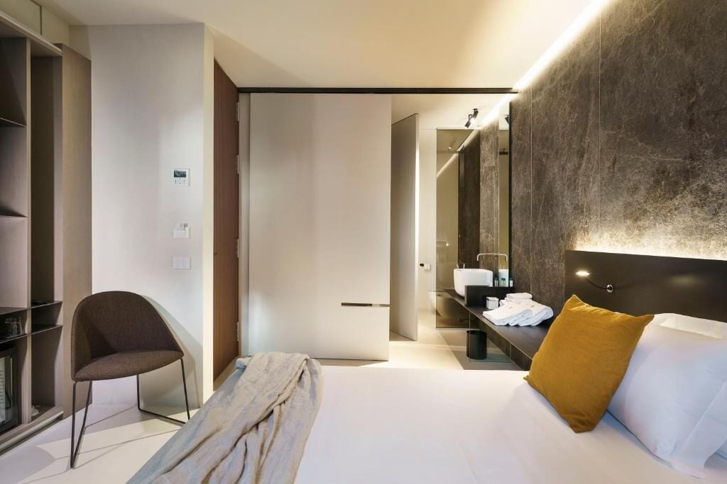 Quarto de hotel moderno com cama, poltrona, guarda-roupa e banheiro visível através de uma porta aberta.