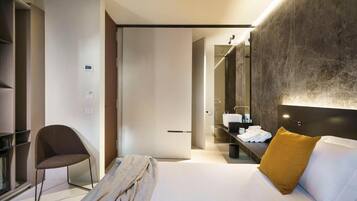 Quarto de hotel moderno com cama, poltrona, guarda-roupa e banheiro visível através de uma porta aberta.
