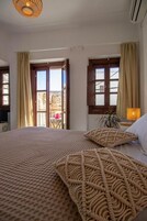 Quarto com uma cama grande, duas janelas com cortinas, uma mesinha de cabeceira com abajur e vista para o exterior.
