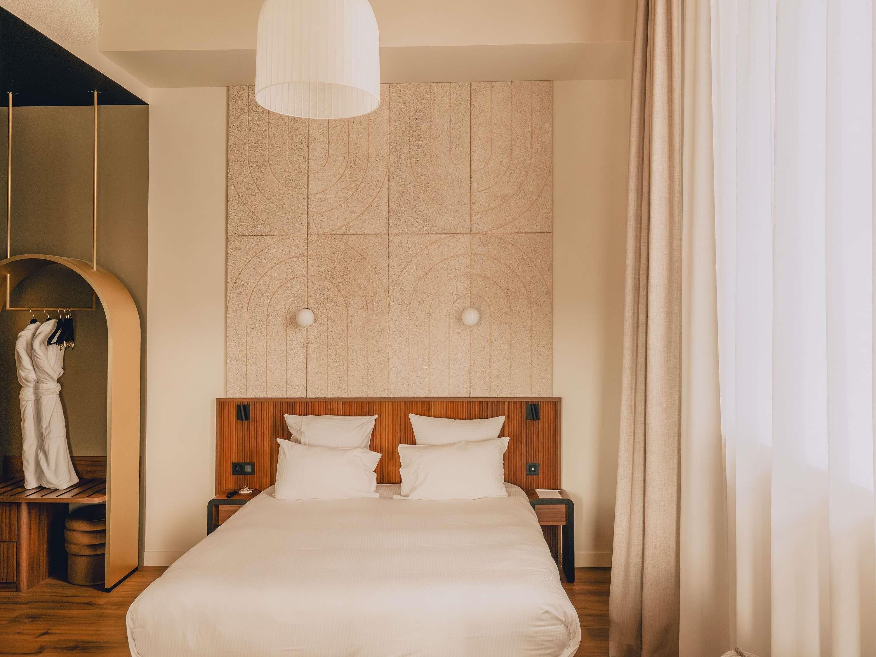 Quarto de hotel com uma cama grande, cabeceira de madeira, duas mesas de cabeceira, luminária pendente e guarda-roupa com espelho.