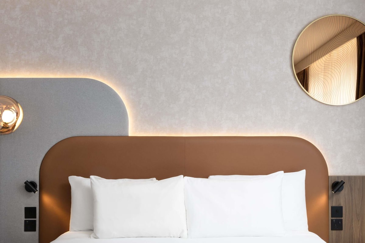 Quarto de hotel moderno com uma cama grande, um espelho redondo e iluminação embutida na parede.