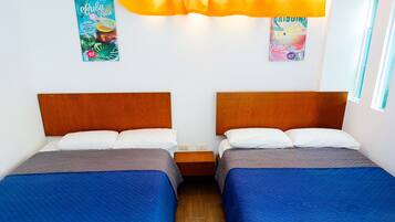 Quarto de hotel com duas camas, cada uma com colcha acolchoada azul e almofadas brancas. Tem cabeceira de madeira, mesa de cabeceira pequena e janela com vista.