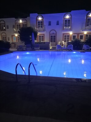 Piscina com superfície de água iluminada em azul, rodeada por espreguiçadeiras e um edifício com janelas e portas em arco.