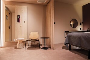 Quarto de hotel com cama, poltrona, mesa pequena, espelho e porta com placa.