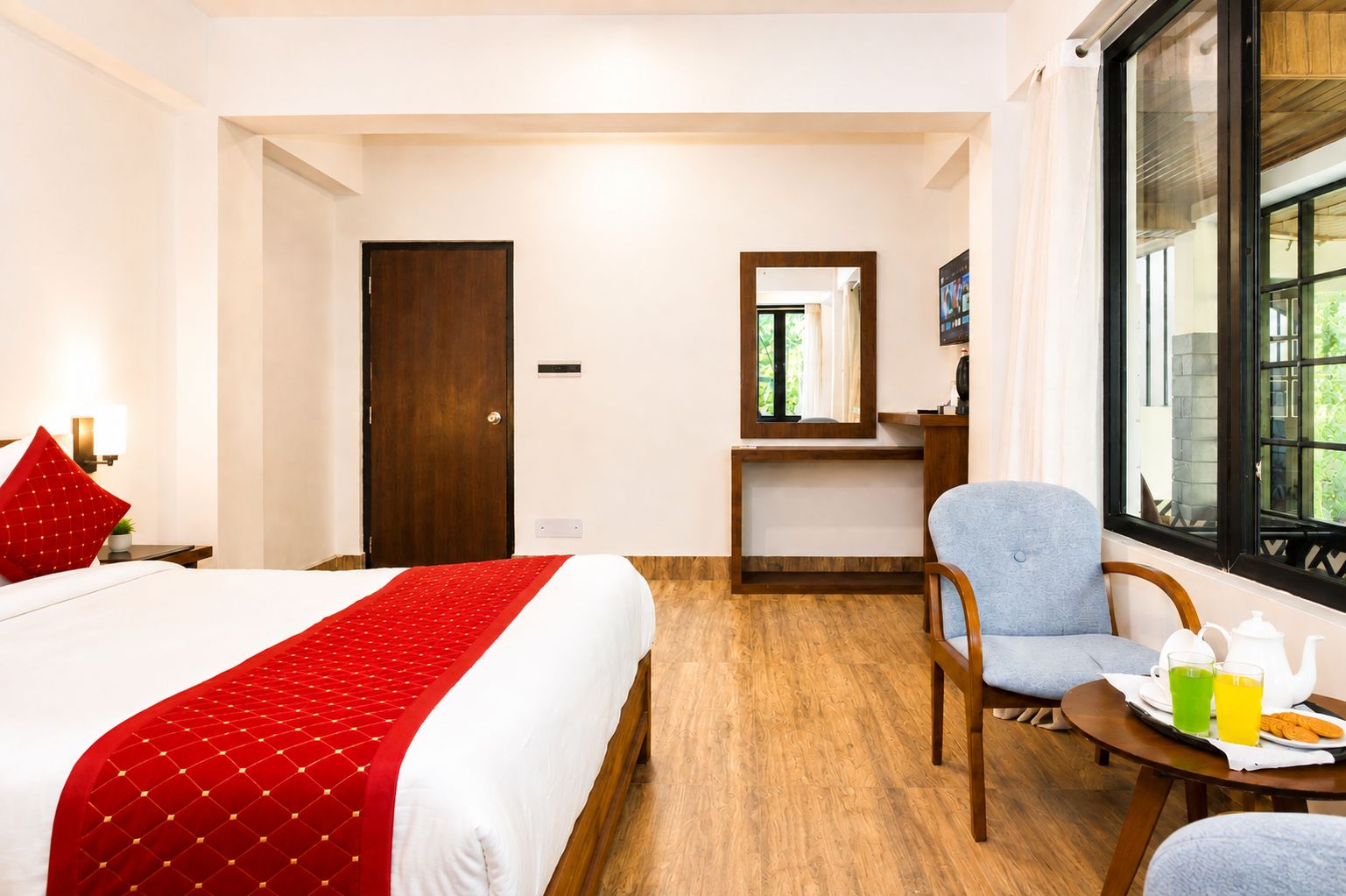 Quarto de hotel com cama, poltrona, mesinha com chaleira e copos, espelho e janela.