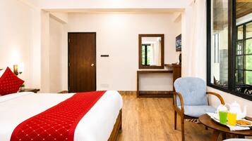 Quarto de hotel com cama, poltrona, mesinha com chaleira e copos, espelho e janela.