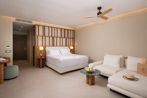 Quarto de hotel moderno com uma cama grande, um sofá, uma mesinha com uma cesta de frutas e um ventilador de teto.