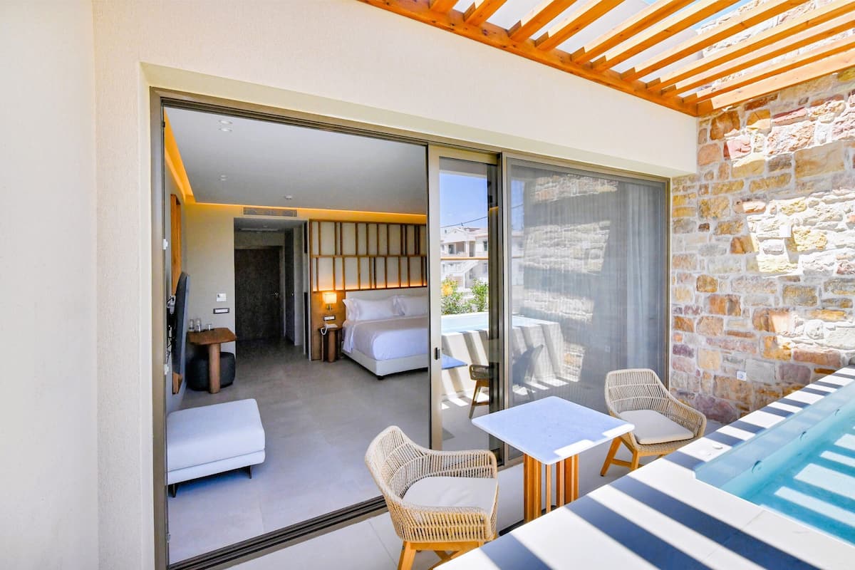Quarto de hotel moderno com porta de vidro para uma varanda, uma pequena mesa, duas cadeiras de vime e área de piscina com muro de pedra.