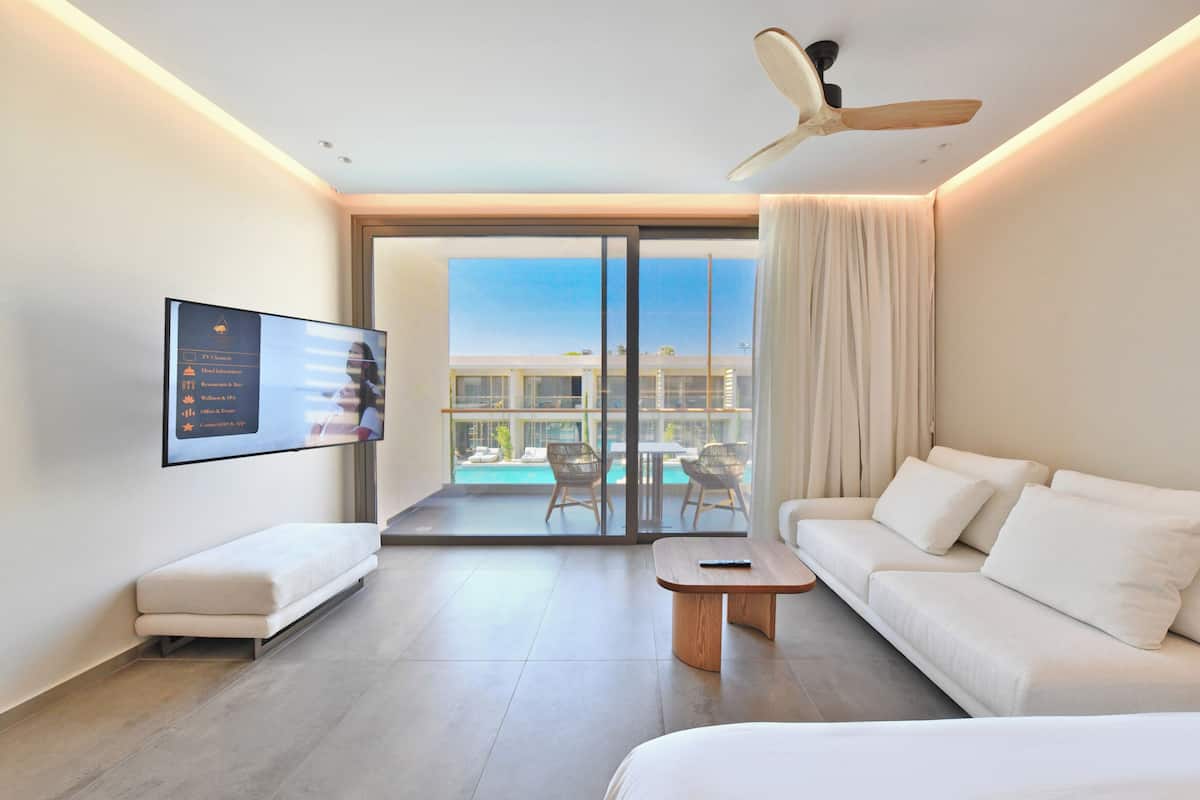 Quarto de hotel moderno com cama grande, sofá, mesa de centro e TV fixada na parede. Tem uma varanda com vista para a piscina.