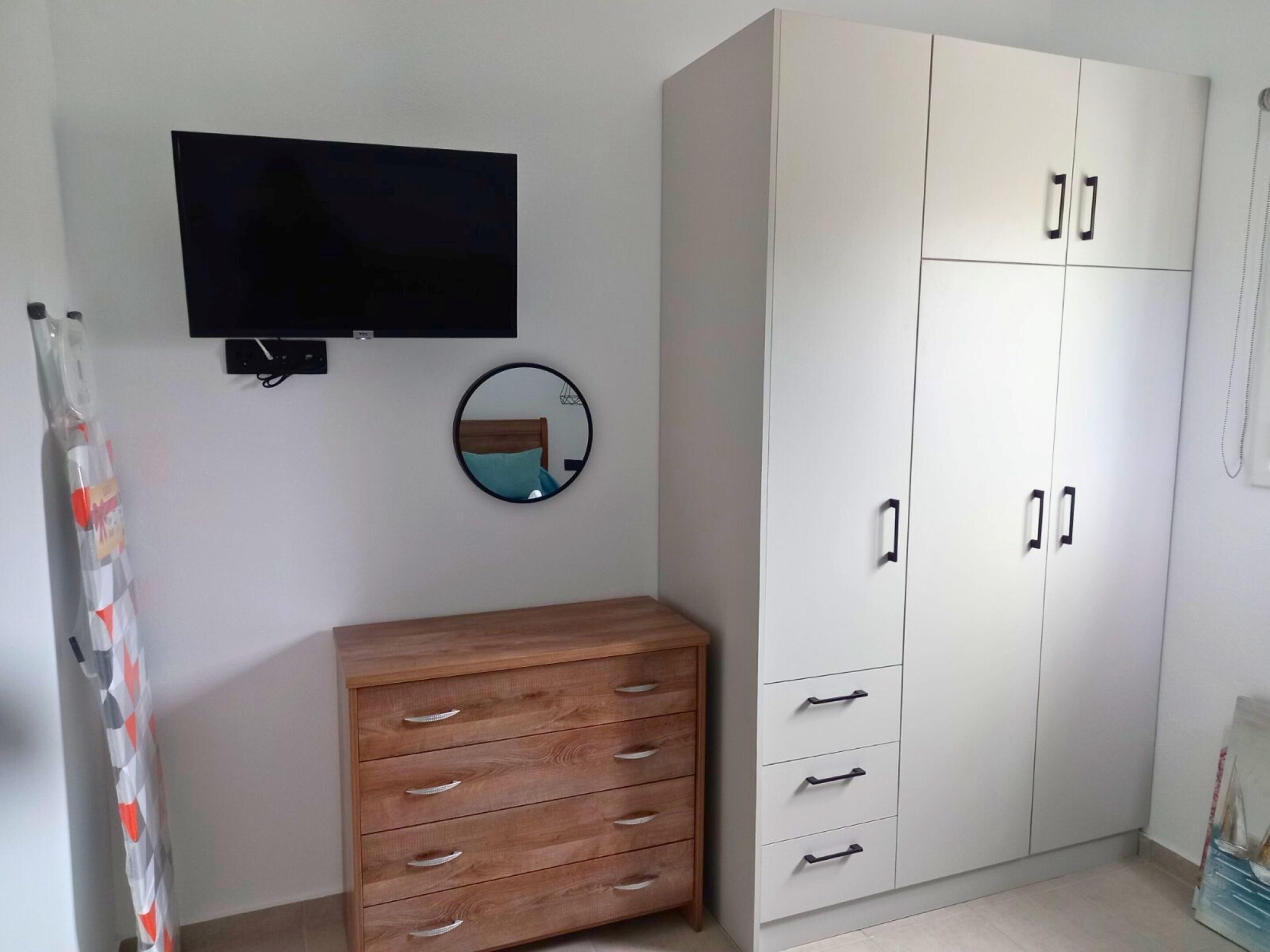Quarto com televisão na parede, cómoda de madeira, guarda-roupa com portas múltiplas e espelho.
