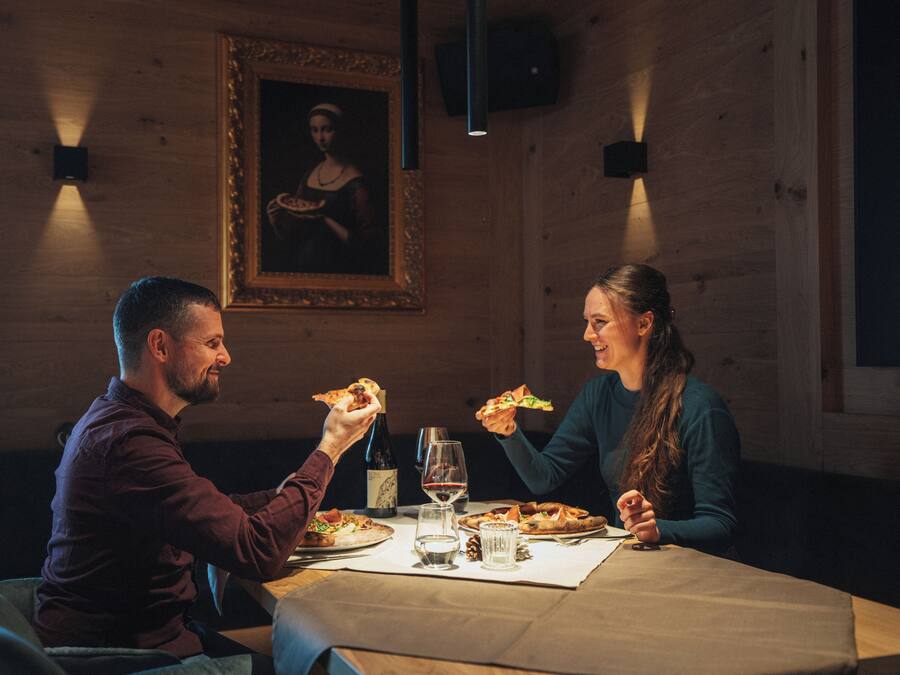 Um casal jantando à mesa, com um quadro na parede, painéis de madeira e uma garrafa de vinho.