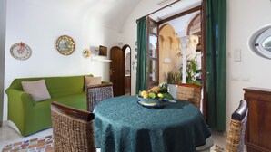 Uma sala de estar com um sofá verde, cadeiras de vime, uma mesa com uma tigela de frutas e vista para uma área externa por uma porta aberta.